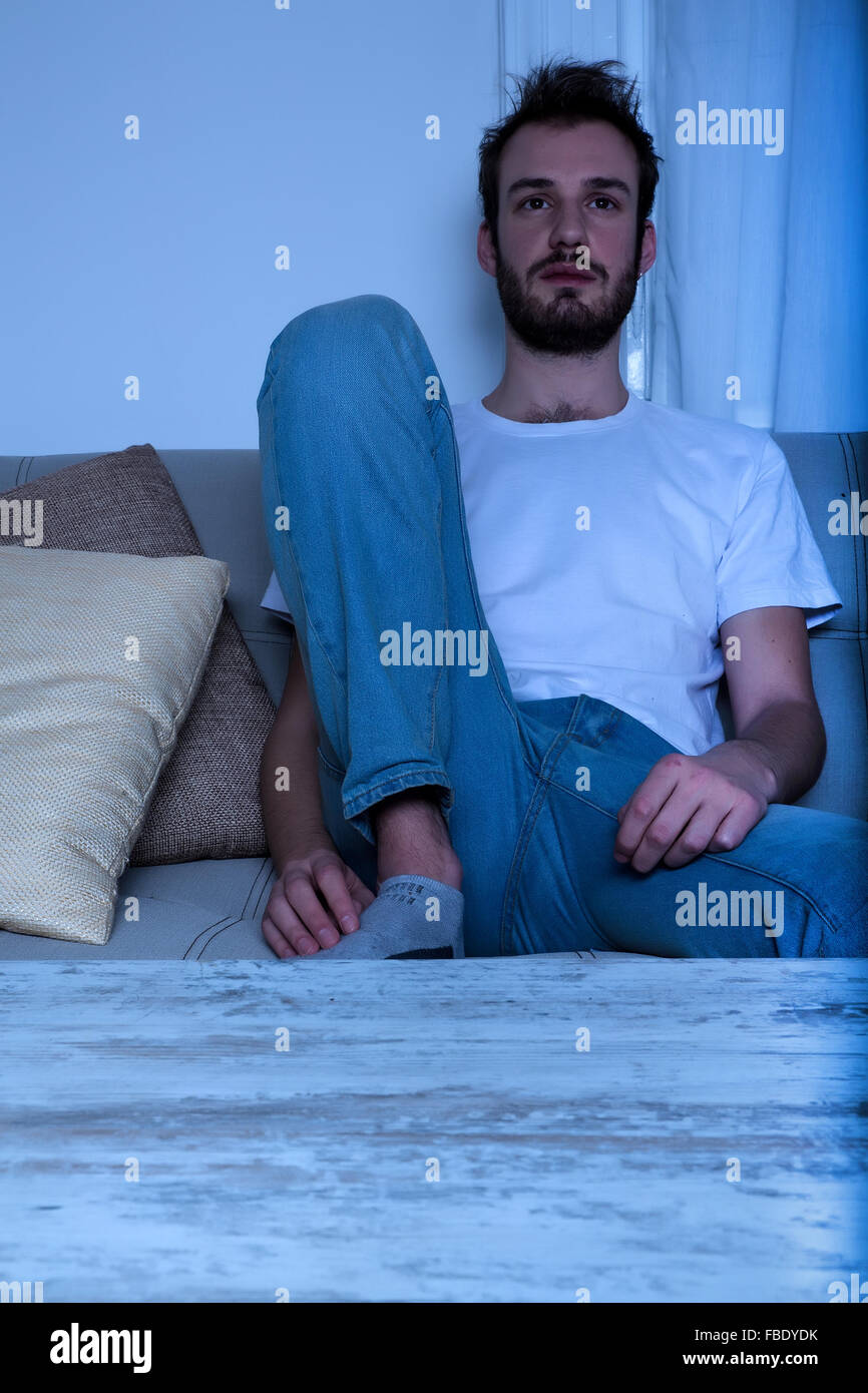 A young bored man sitting on the sofa in front of the TV at night while ...