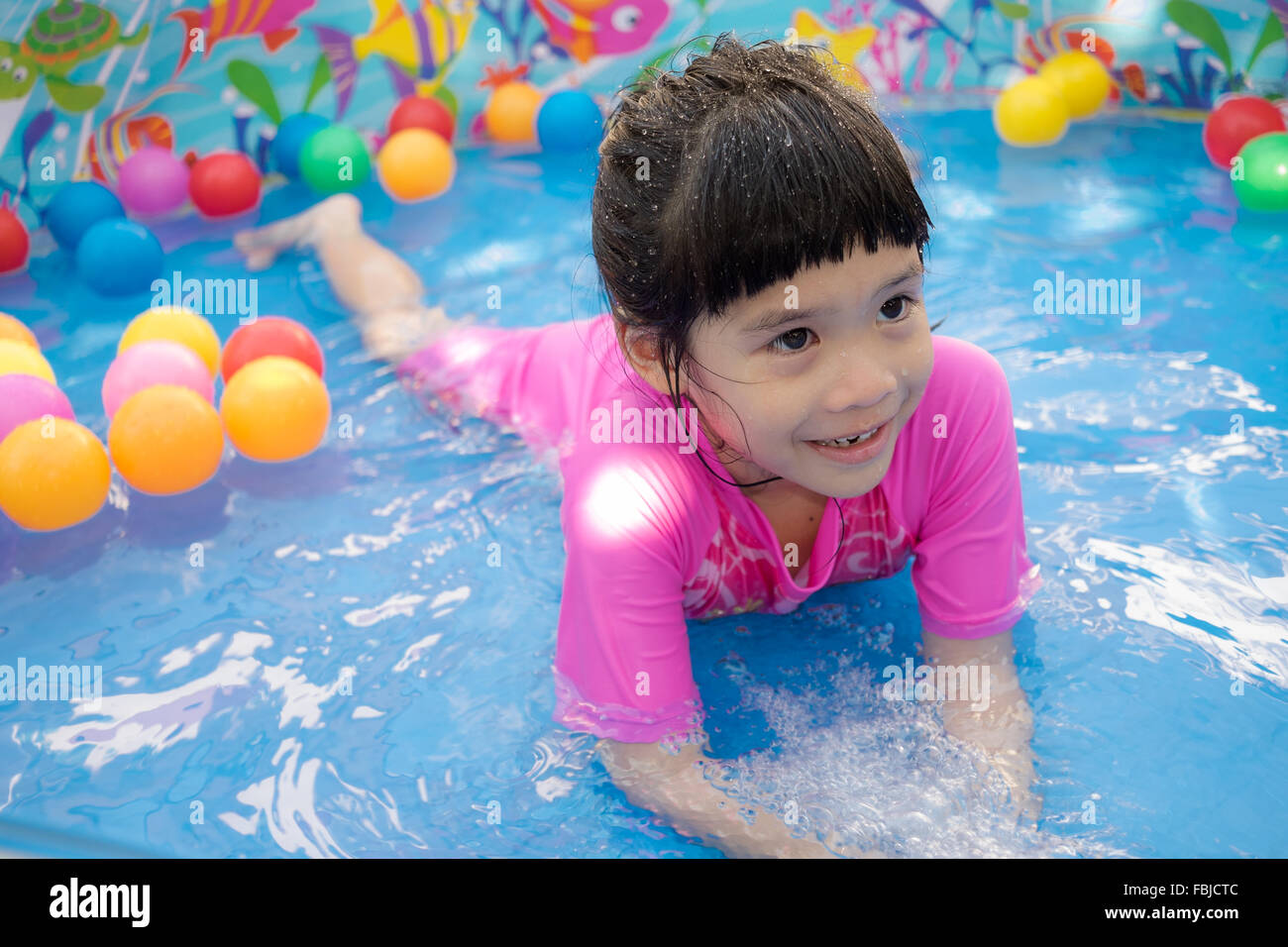A baby girl in pink suit playing water and balls in blue kiddie pool ...