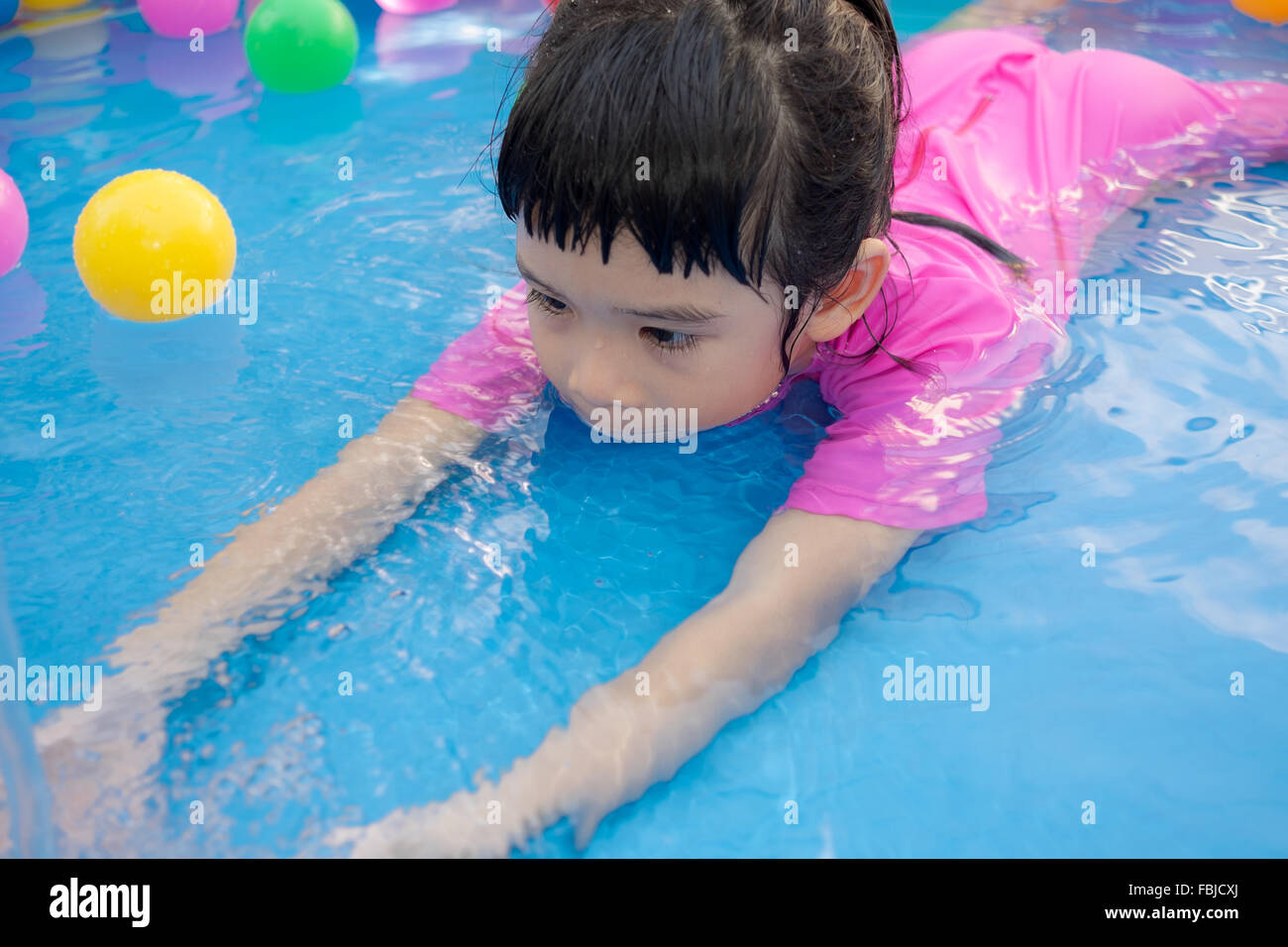 A baby girl in pink suit playing water and balls in blue kiddie pool ...