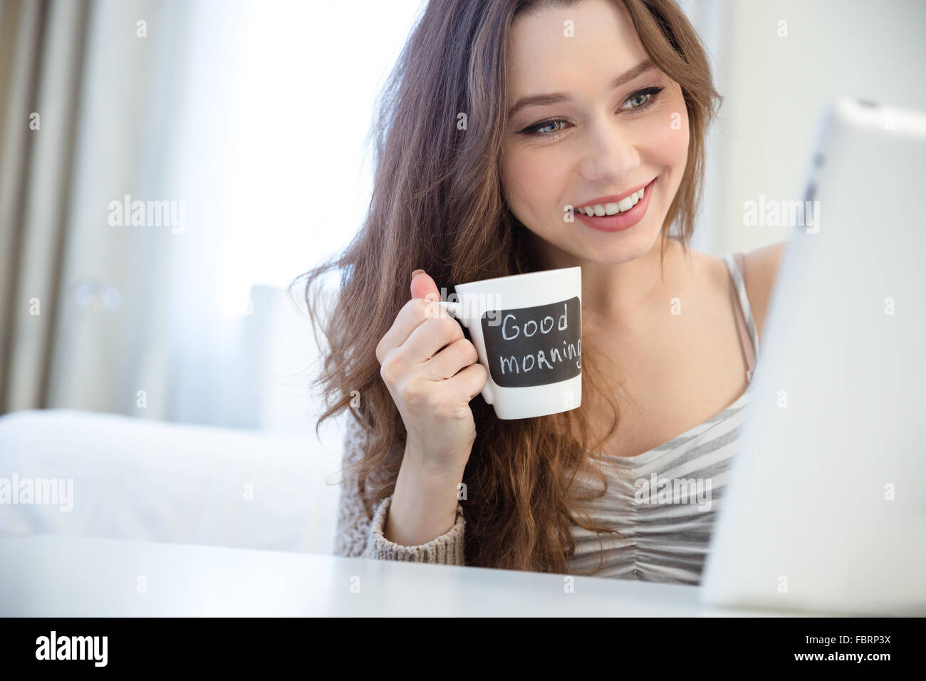 Smiling charming young woman using tablet and drinking coffee from ...