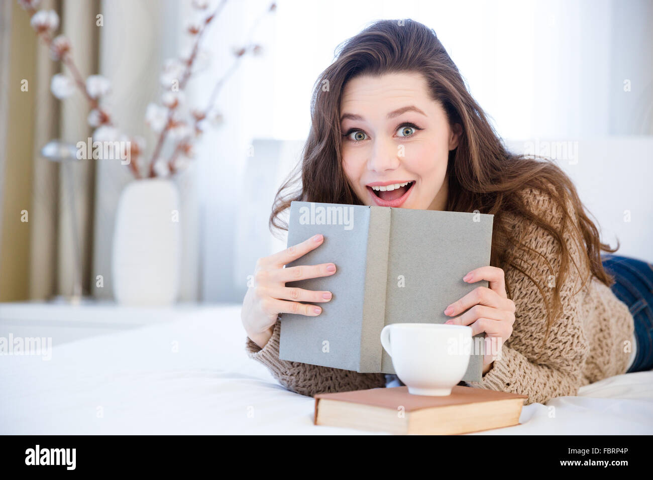 Happy amazed charming young woman lying on bed and reading book in ...