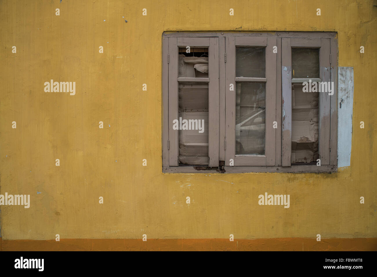 A roughly painted yellow wall with an off centre window frame. Broken ...