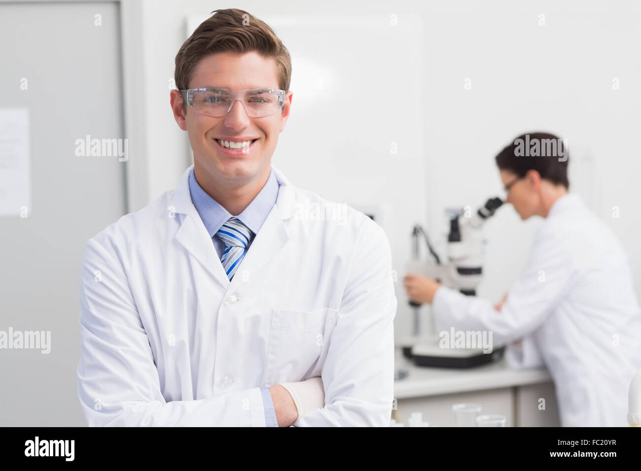 Scientist smiling at camera arms crossed and another working with ...