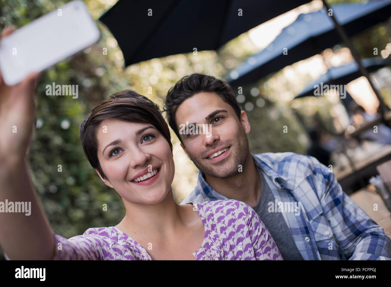 A couple seated at an outdoor city cafe, taking a selfy with a smart ...