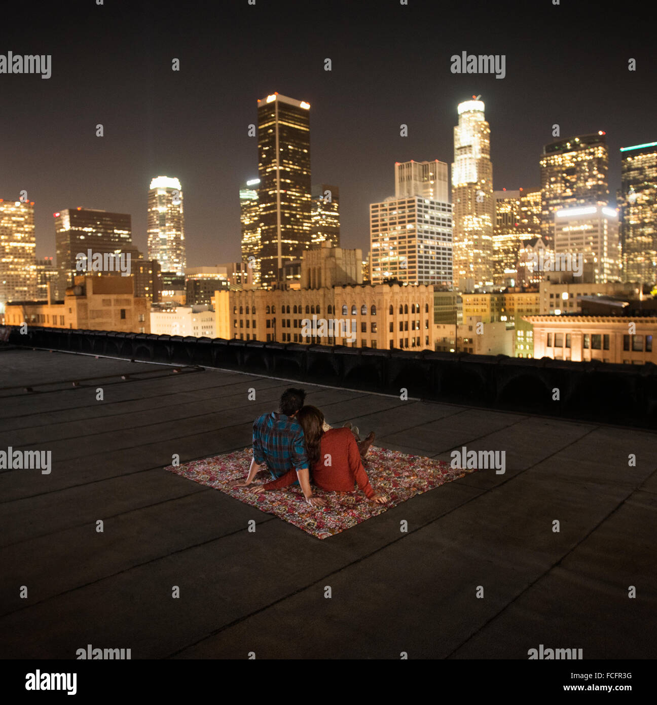 A couple sitting on a rug on a rooftop overlooking a city lit up at ...
