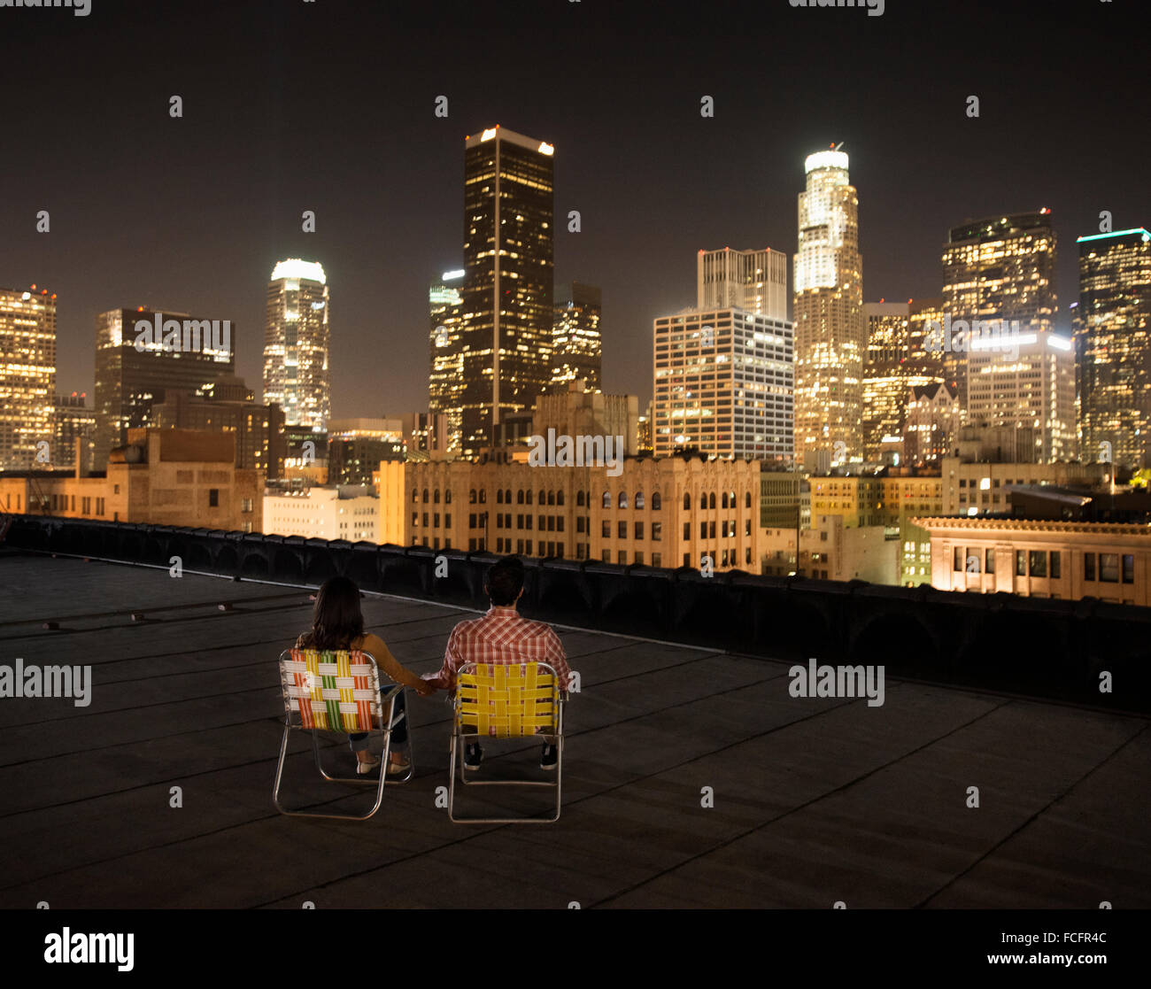 A couple on a rooftop overlooking Los Angeles at night, sitting side by ...