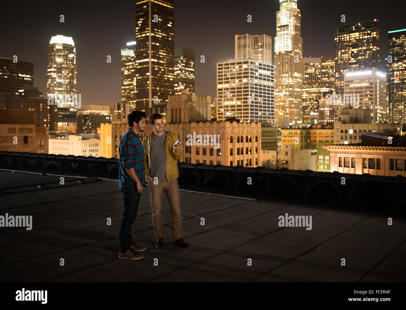 Two men on a rooftop overlooking Los Angeles at night, looking at a ...