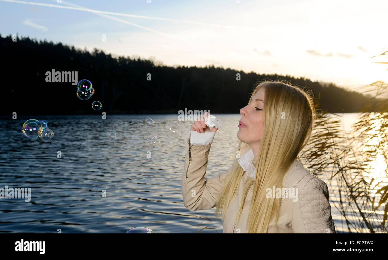 Blond young woman blowing soap bubbles in front of a lake Stock Photo ...