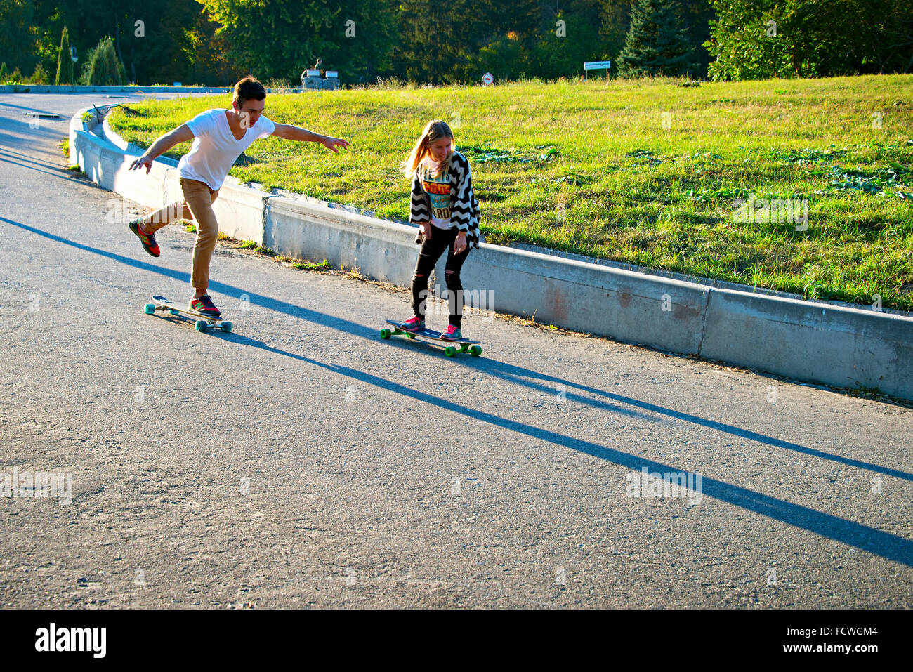 Young couple riding the longboards down the hill at sunset in a park ...