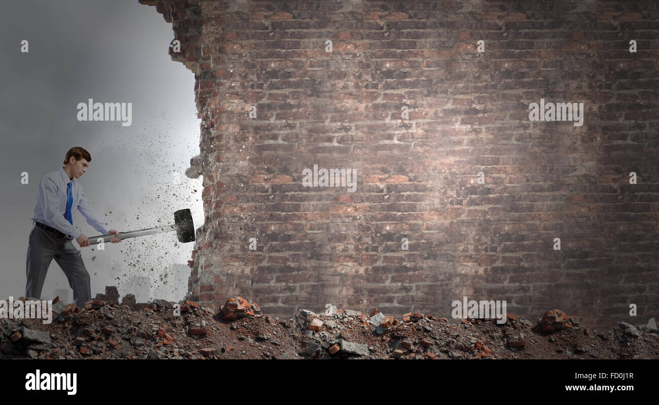 Young determined businessman with big hammer in hands crashing wall ...