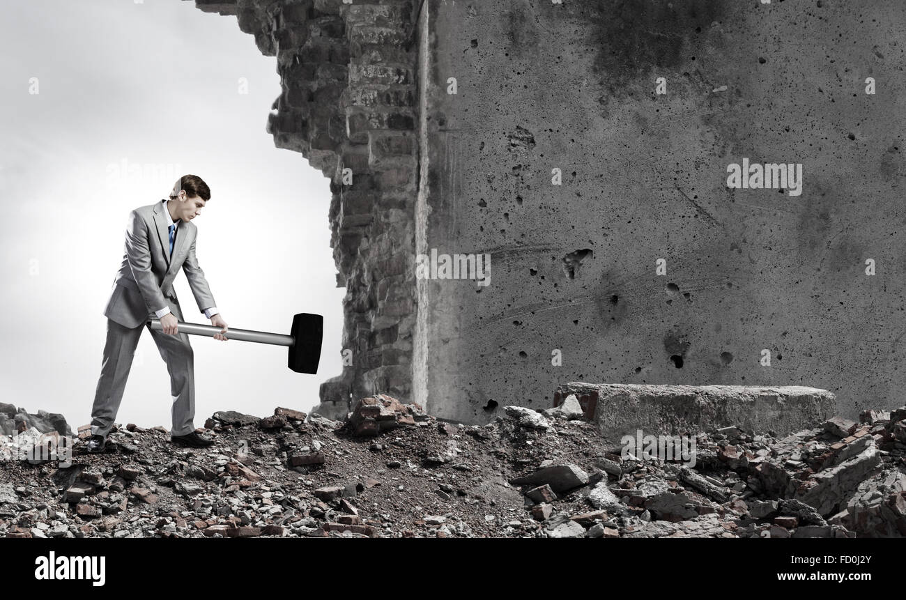 Young determined businessman with big hammer in hands crashing wall ...
