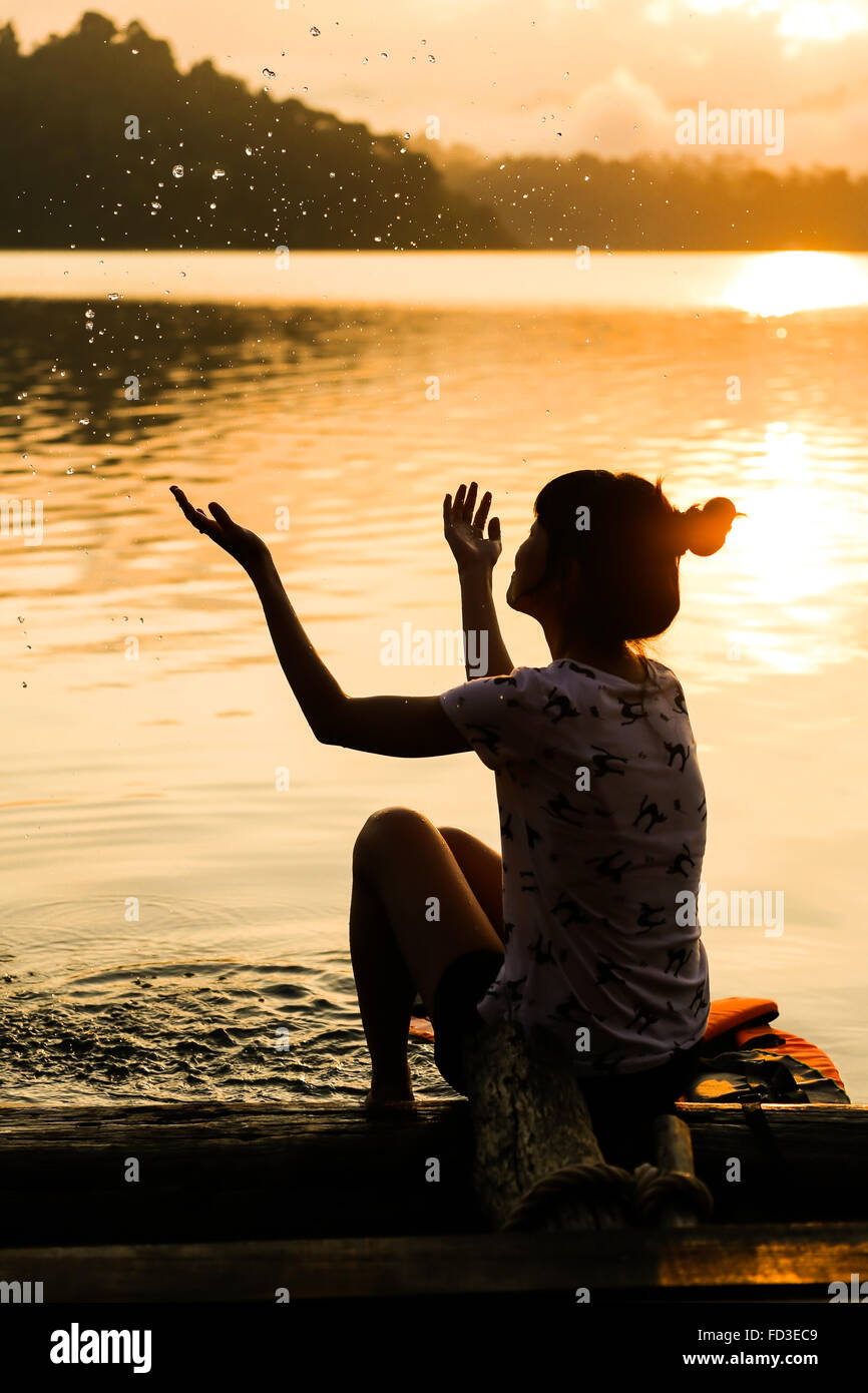 Silhouetted Thai girl sitting on a wood raft is splashing water in the ...