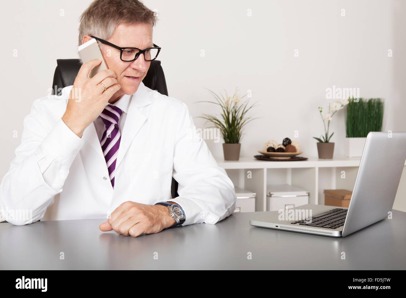 Doctor chatting on his mobile phone while seated at his desk in his ...