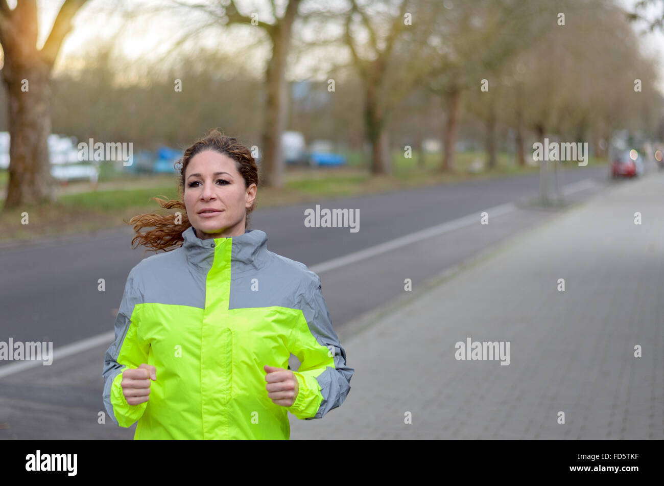 Determined woman jogging along a street in a high visibility jacket ...