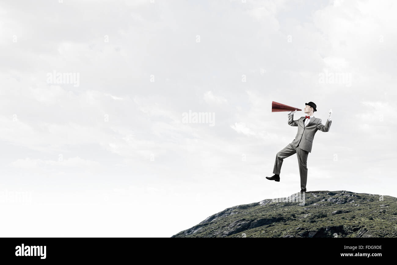 Young businessman on rock top screaming emotionally in paper trumpet ...