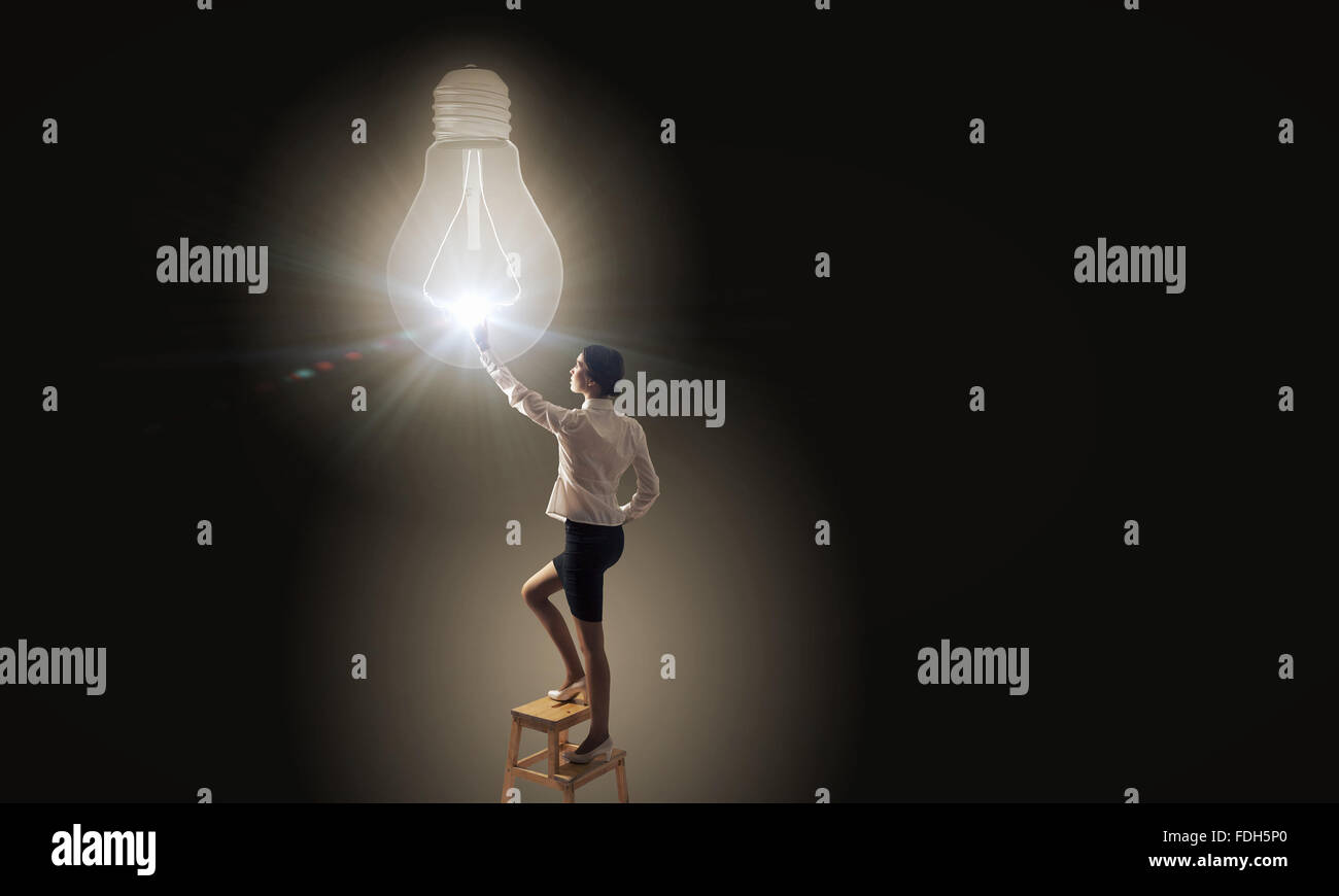 Back view of businesswoman standing on chair and reaching light bulb ...
