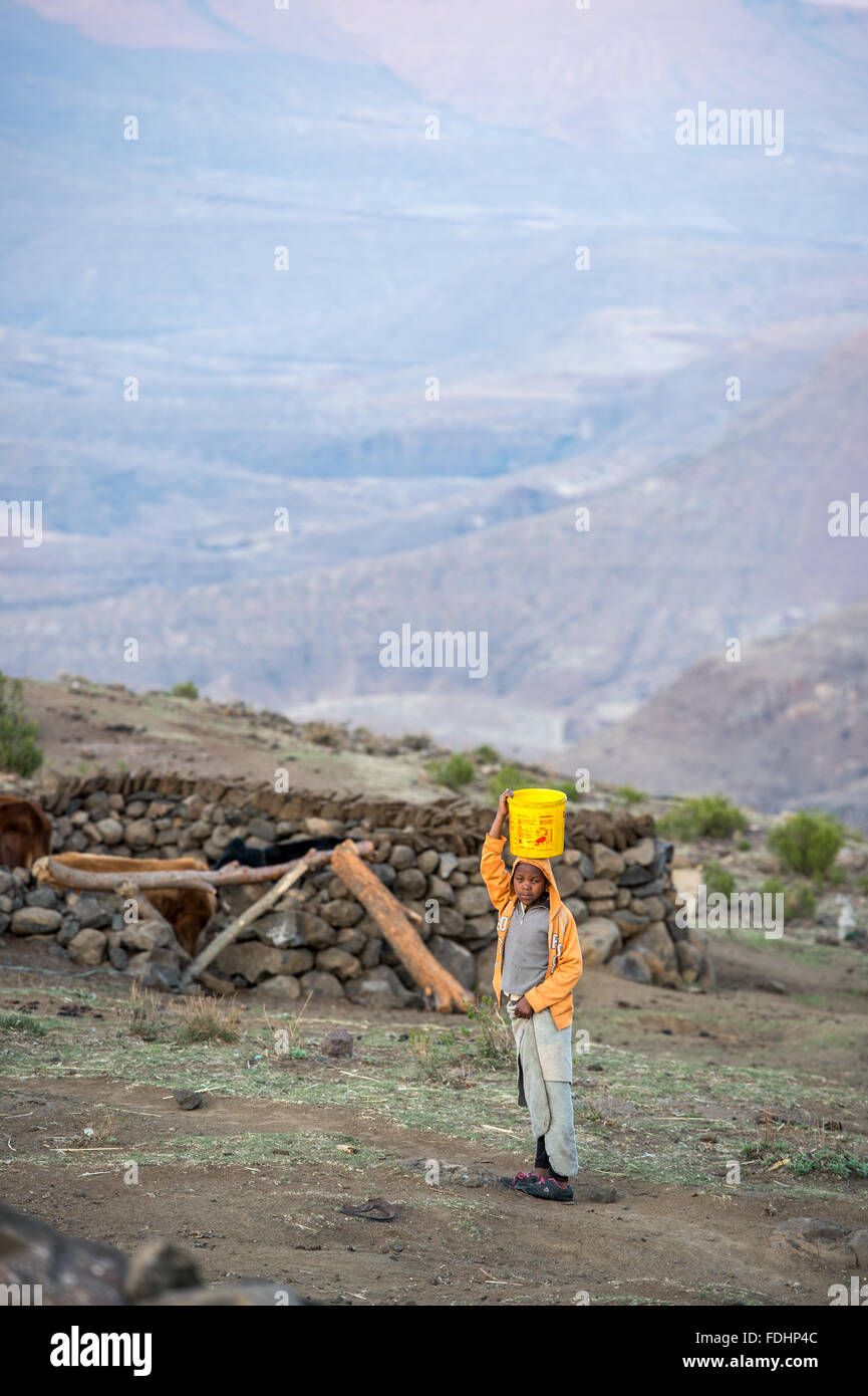 Young boy carrying a bucket on his head in front of a mountain range in ...