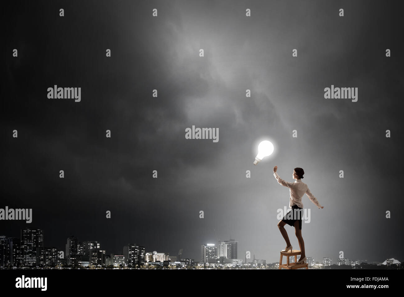 Back view of businesswoman standing on chair and reaching light bulb ...