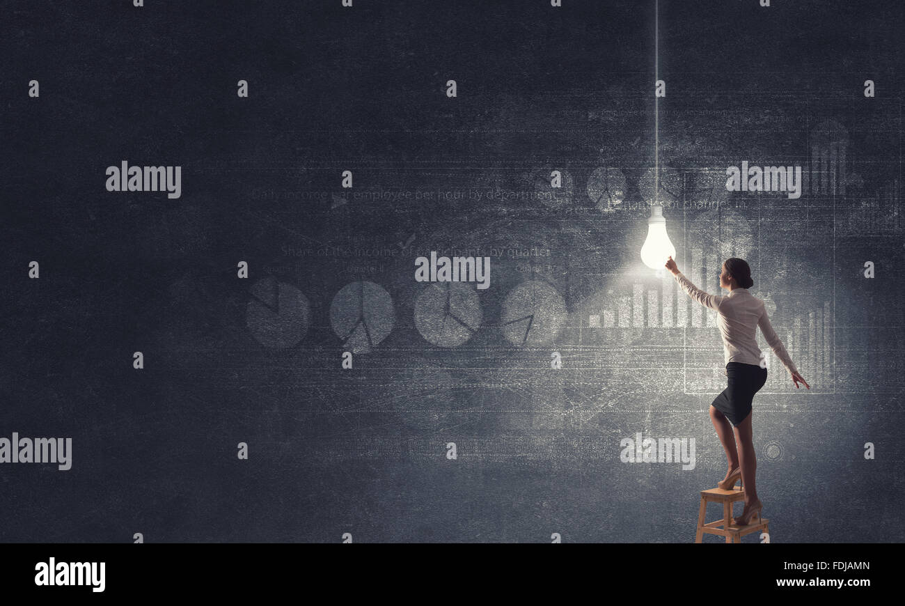 Back view of businesswoman standing on chair and reaching light bulb ...