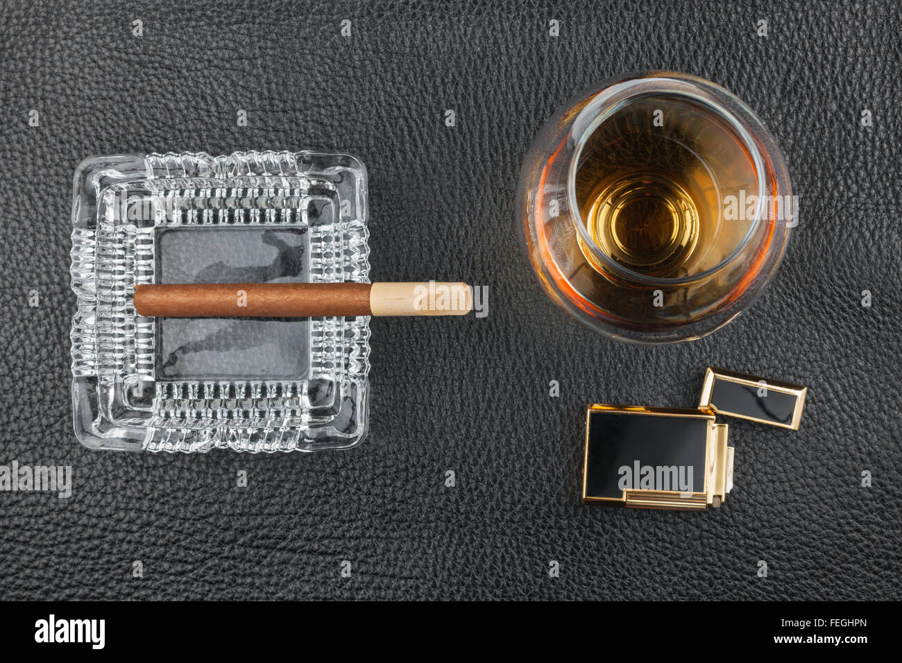 Golden lighter with a glass of alcohol and cigar lying on a black skin ...