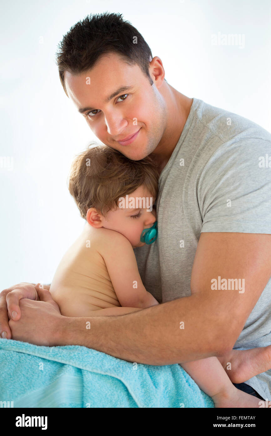 Sleepy baby boy cuddling his father, who is smiling at the camera with ...