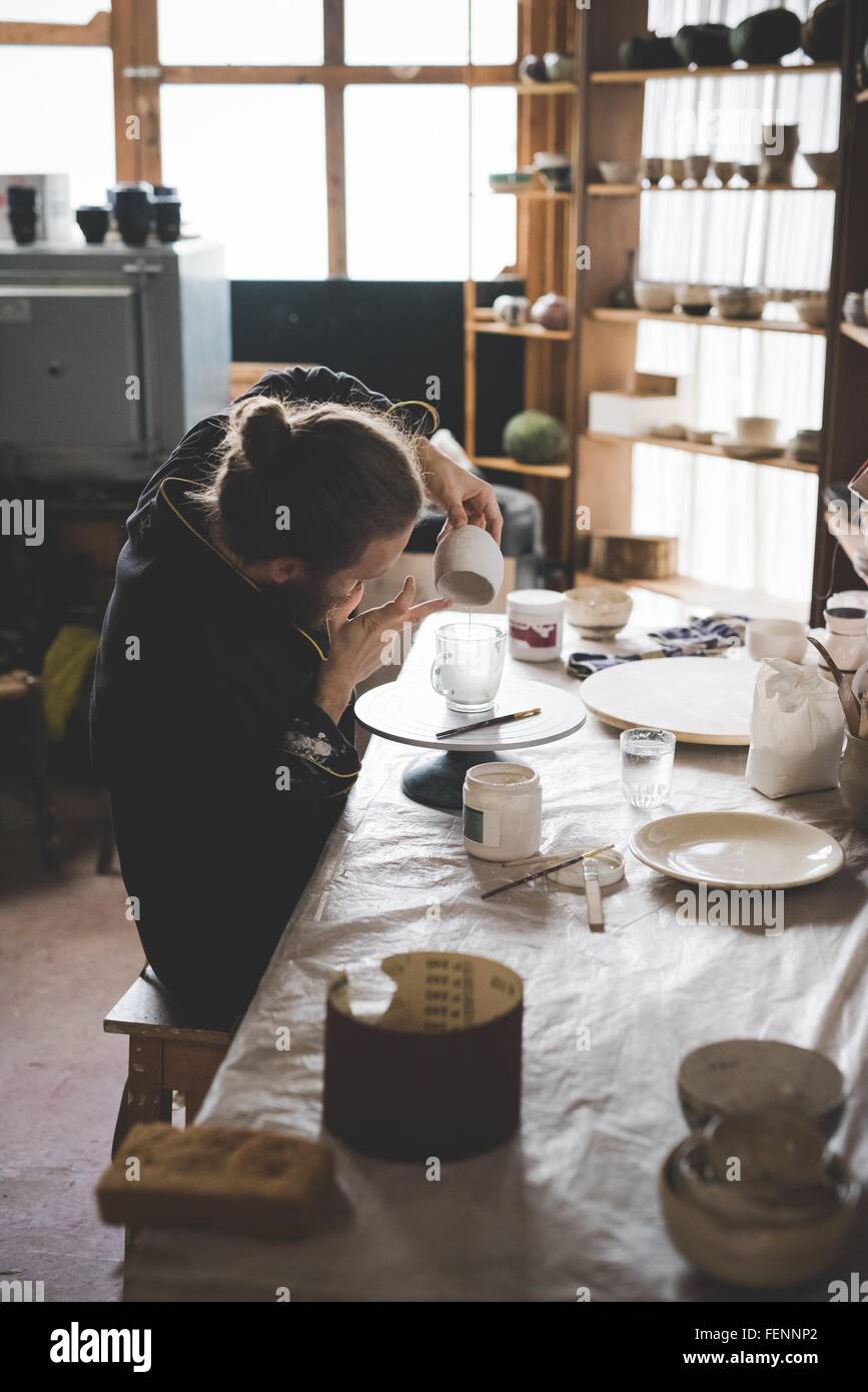 Side view of mid adult man sitting in workshop applying ceramic glaze ...