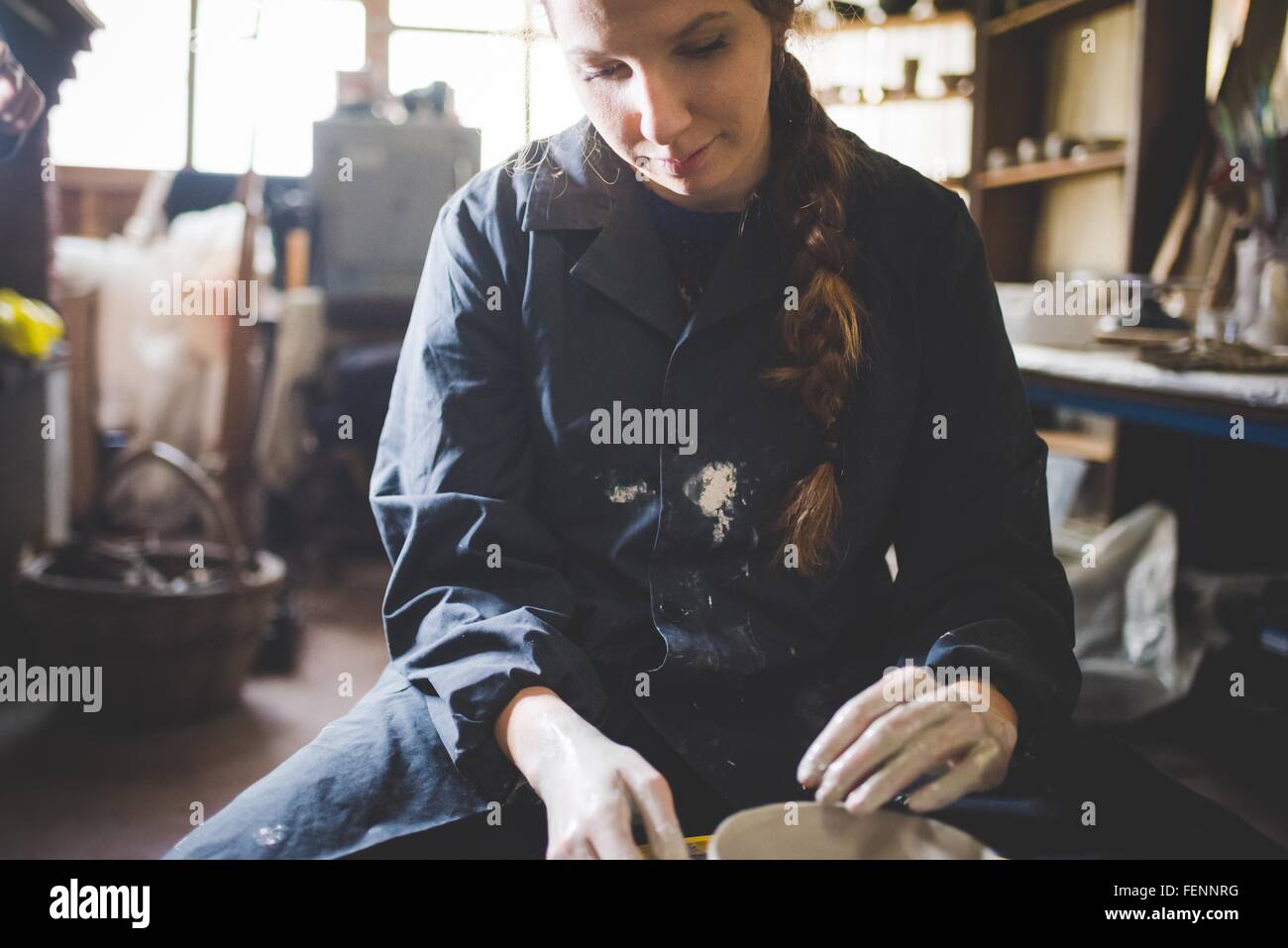 Front view of young woman sitting at pottery wheel looking down making ...