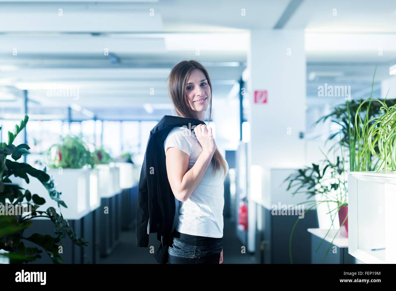 Side view of young woman in office carrying jacket over shoulder ...
