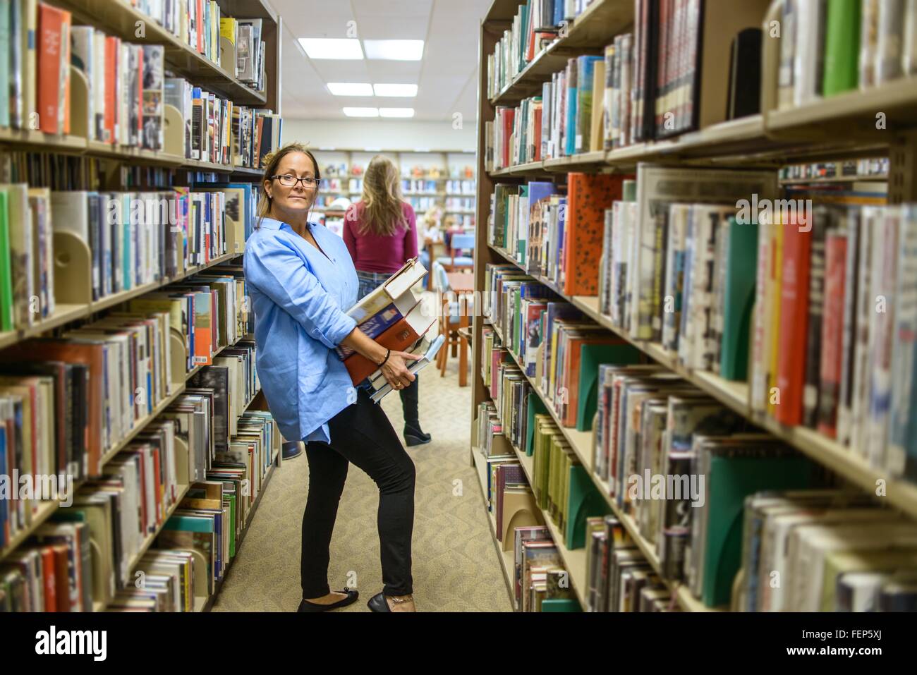 Portrait of mature woman carrying stack of books from library shelves ...