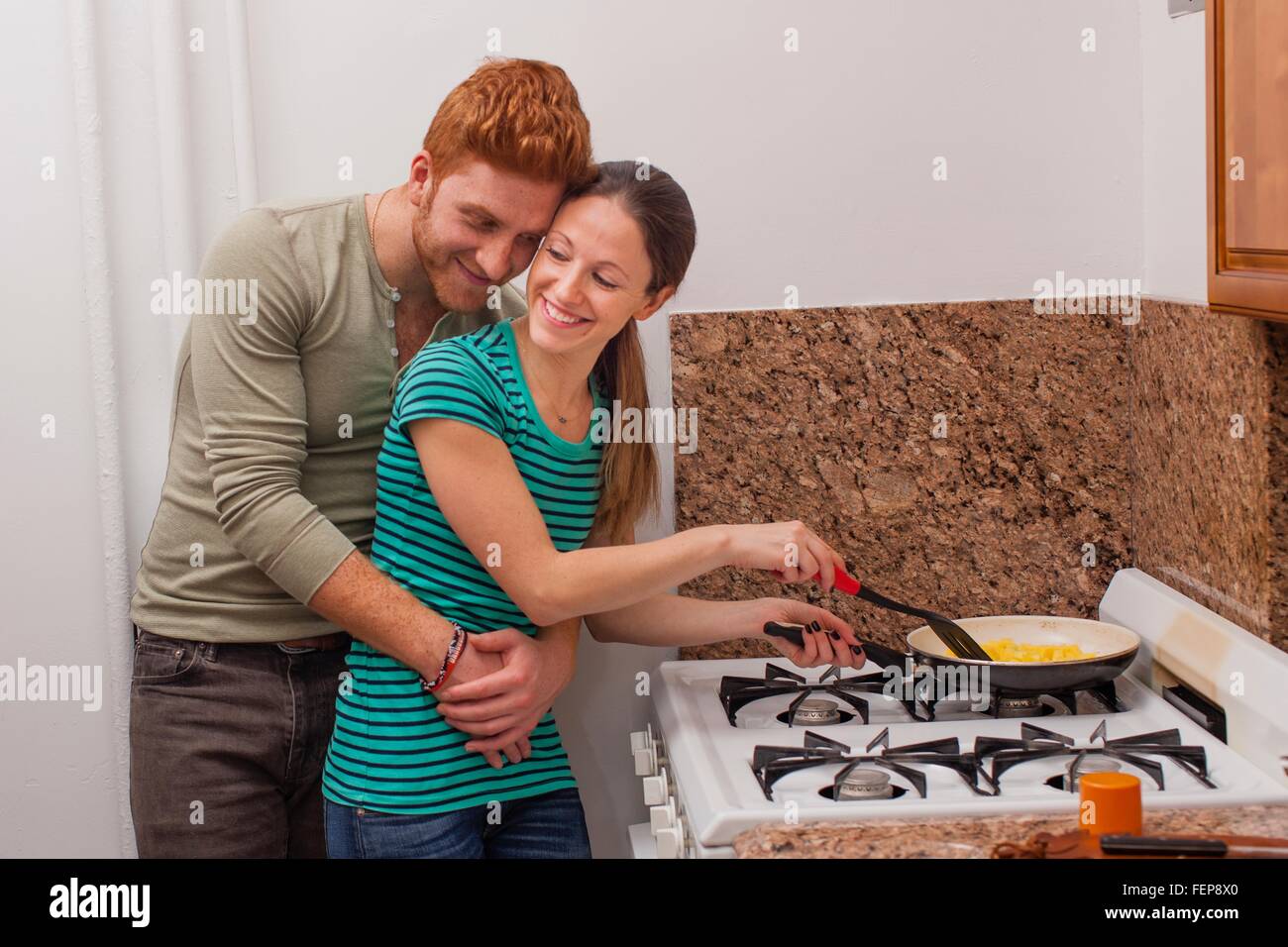 Young man in kitchen arms around young woman cooking on hob smiling ...