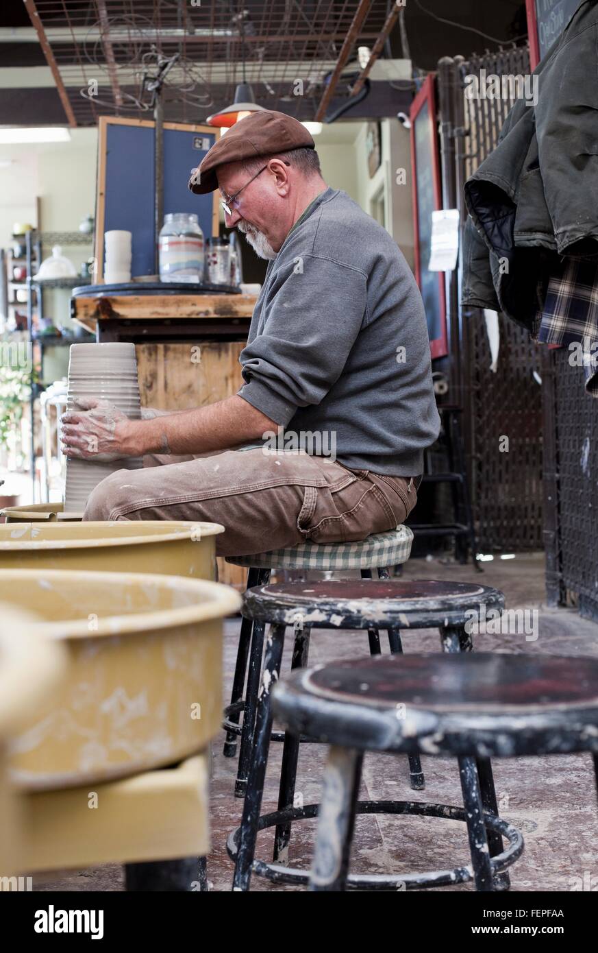 Side view of potter in workshop sitting at pottery wheel shaping clay ...