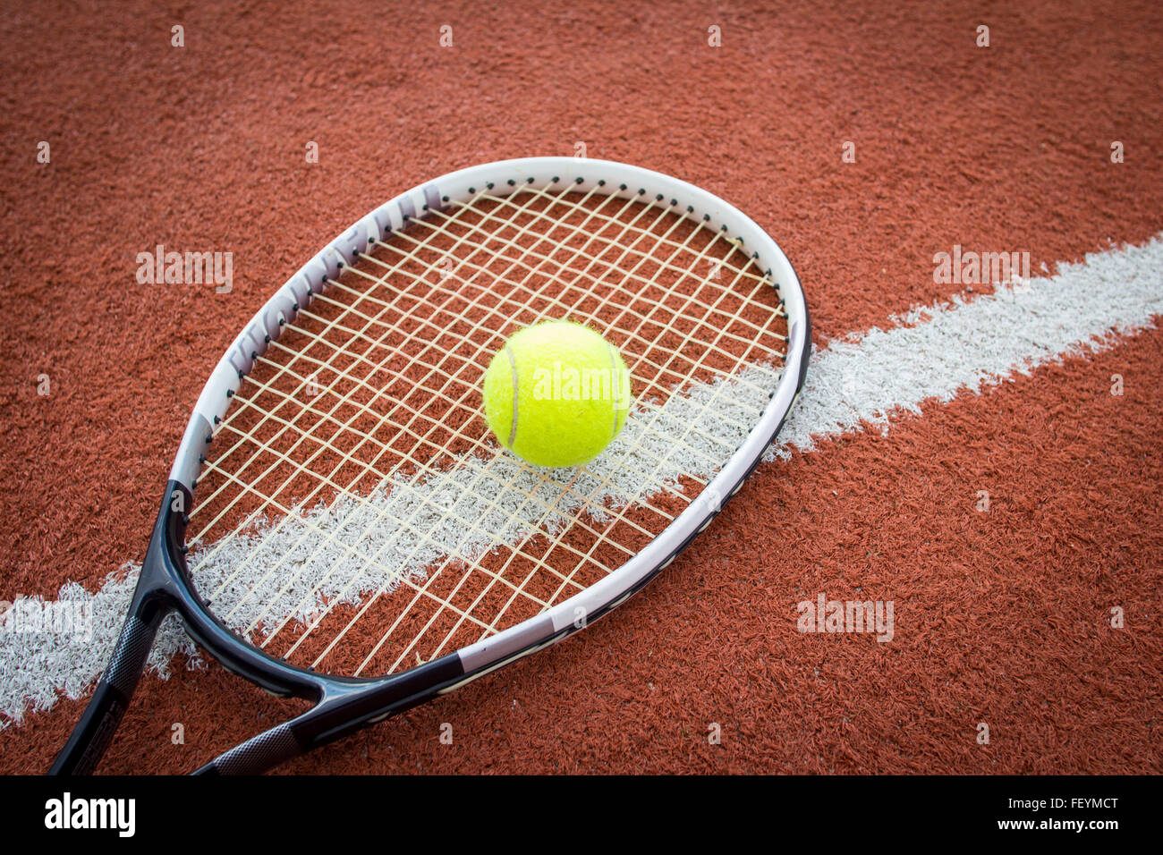 A black and white tennis racket near a yellow ball on a brick red court ...