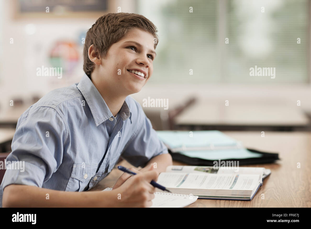 A boy sitting at a desk in class using school books and holding a pen ...