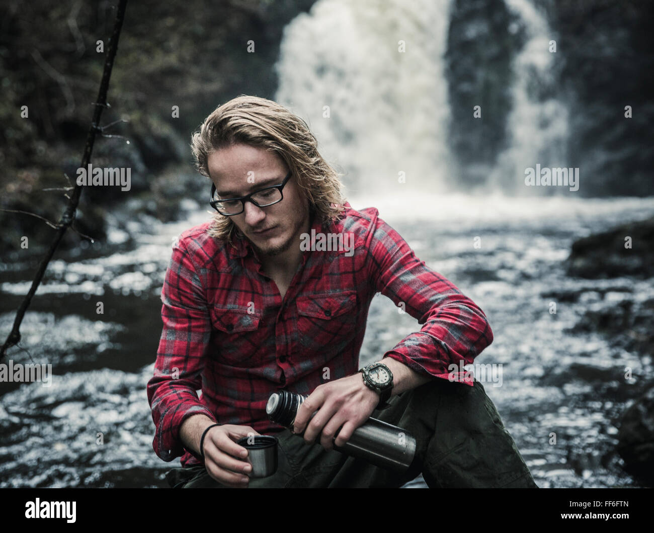A young man sitting pouring a hot drink from a flask by a fast flowing ...