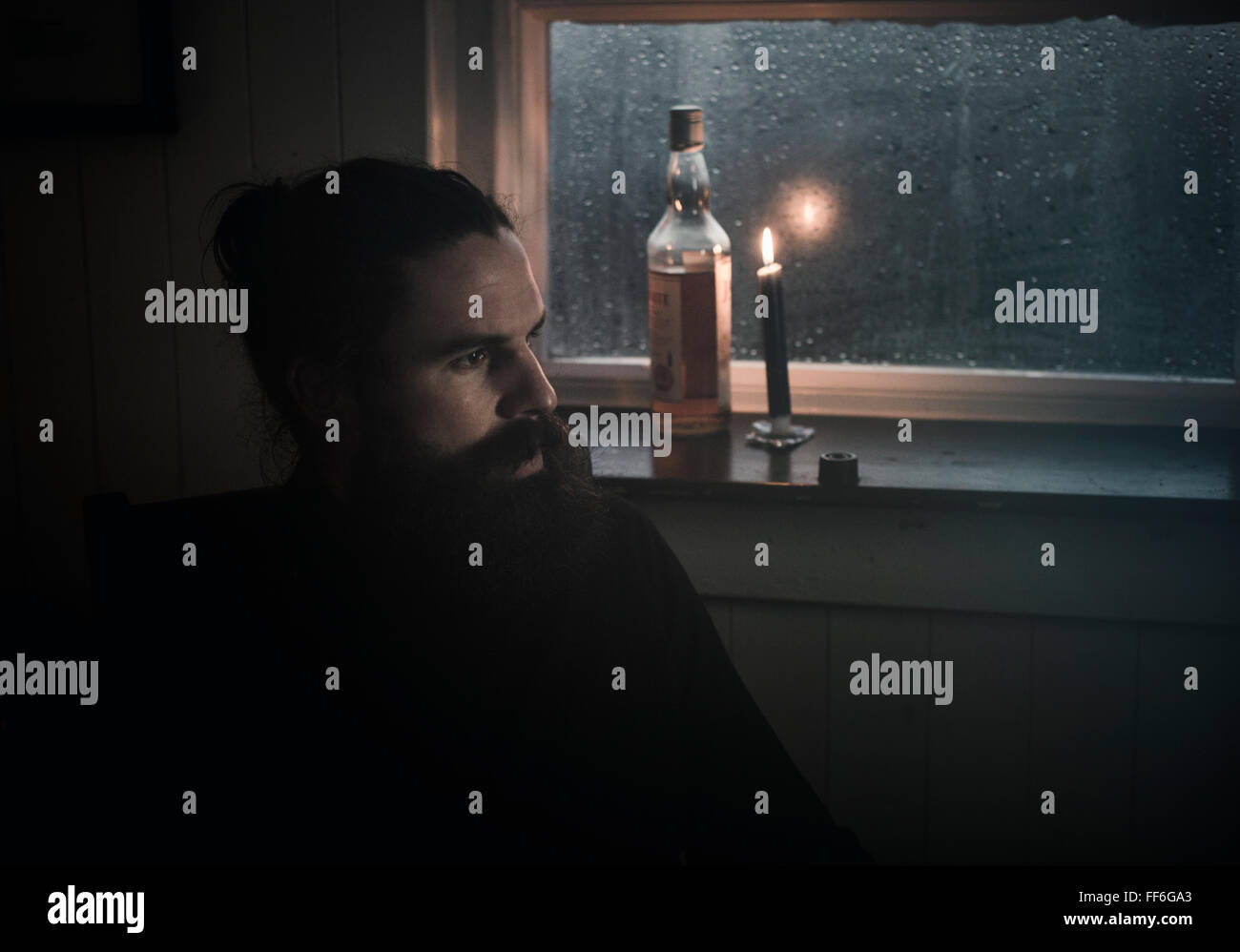 A man sitting in the dark by a window in candlelight drinking from a ...