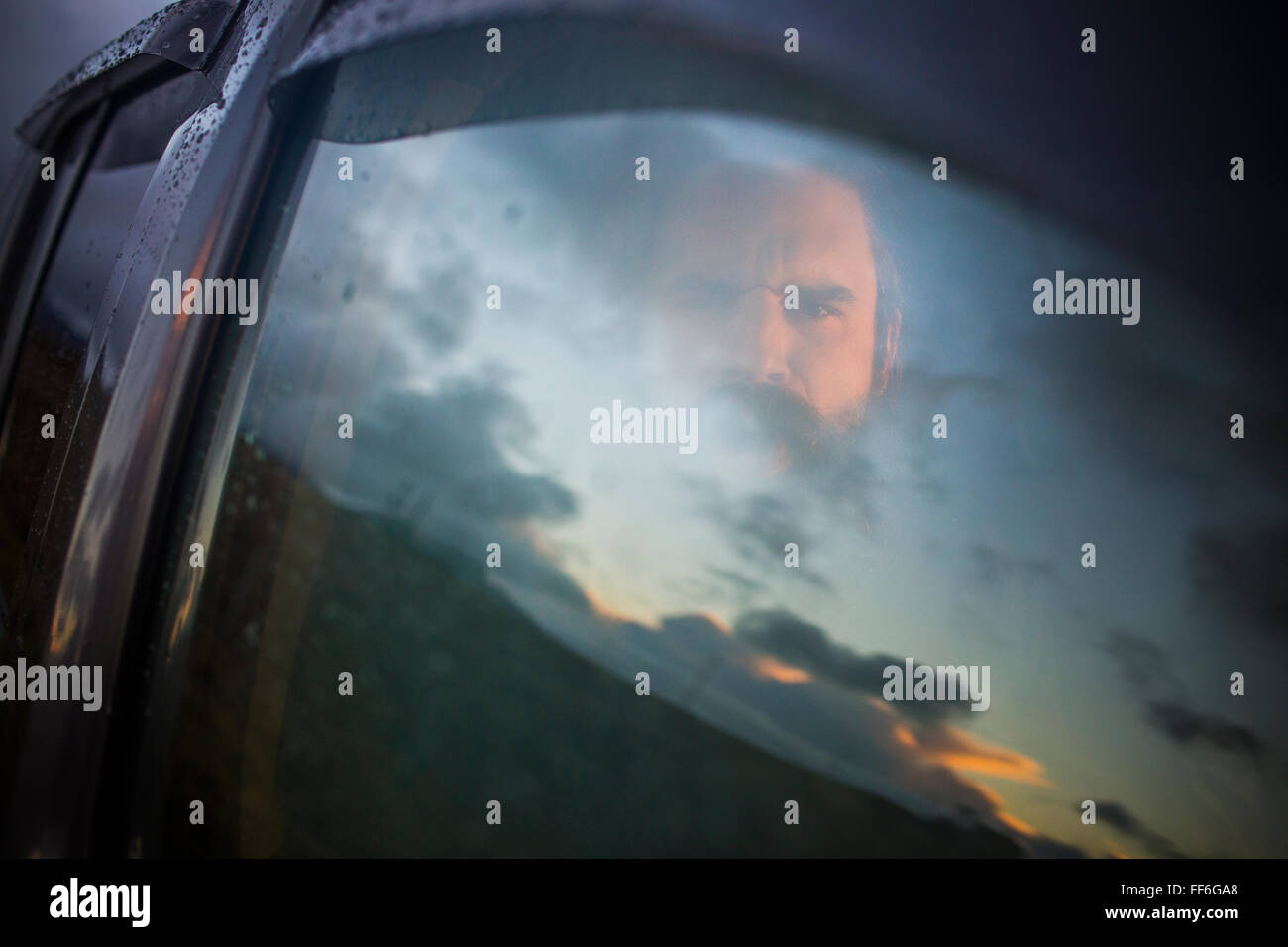 A man sitting in a car looking out. Reflections of the sunset sky on ...