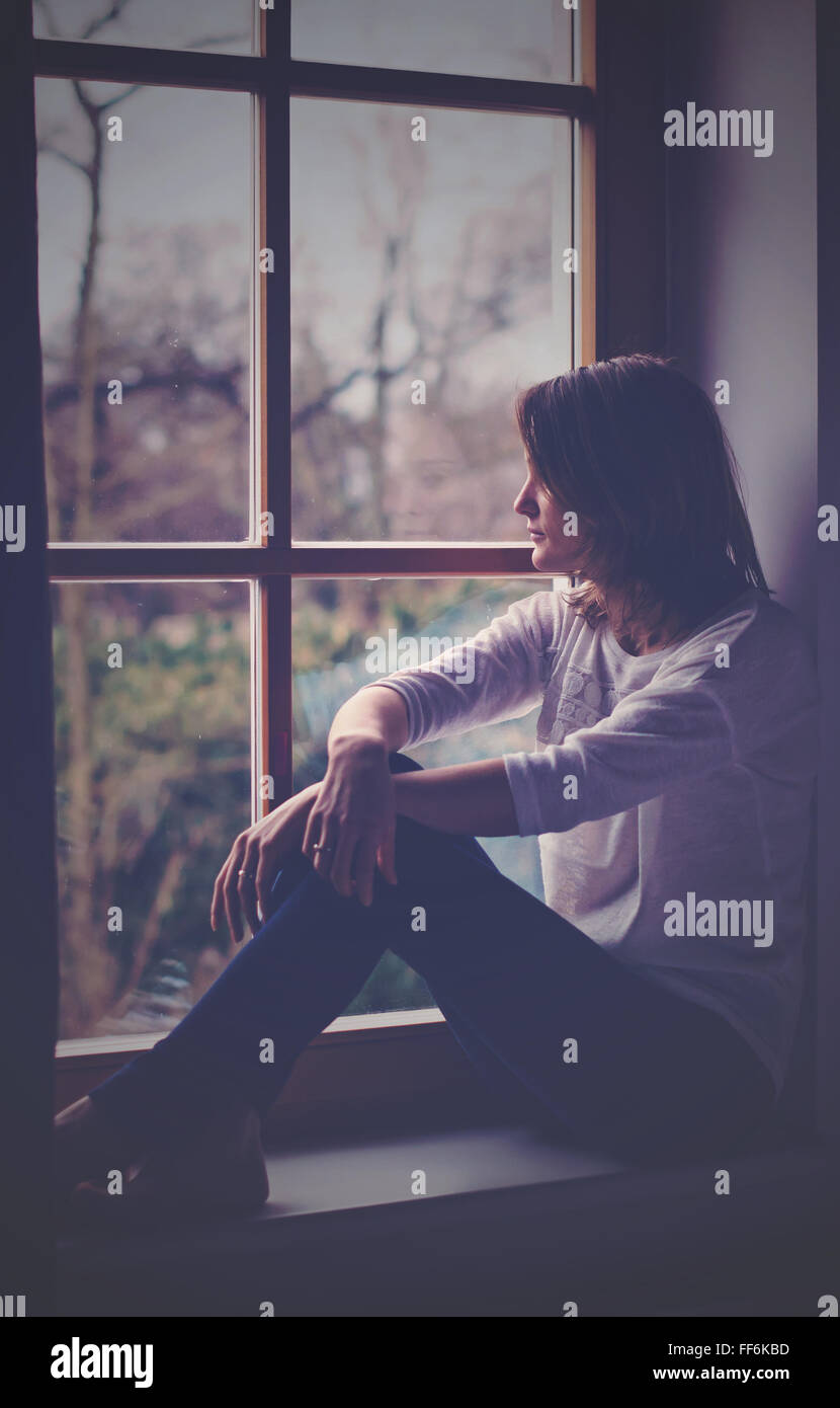 Young attractive woman, sitting on a window, looking outside, lonely ...