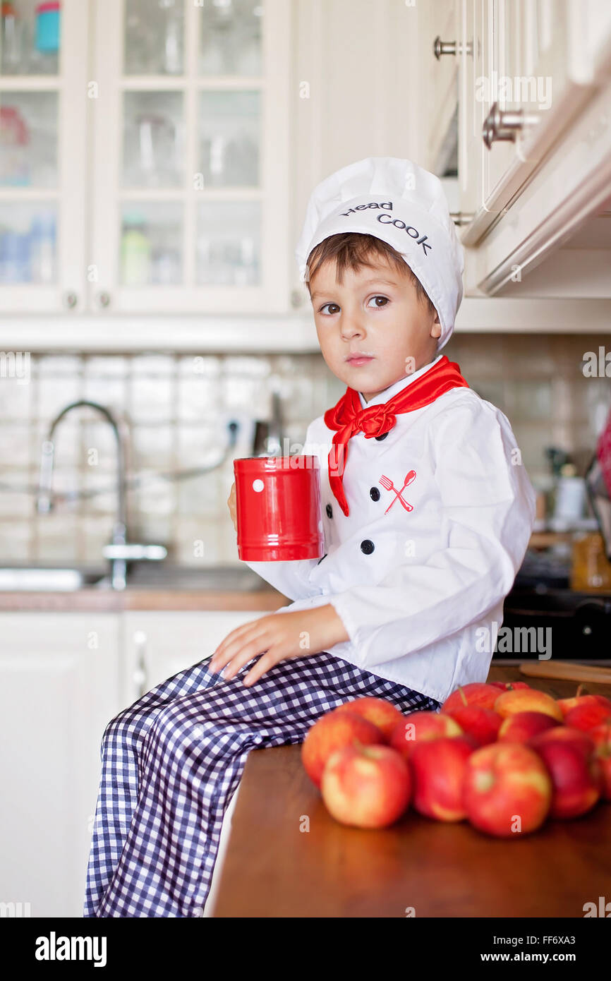 Sweet little boy, dressed as a chef, eating and cutting apples for pie ...