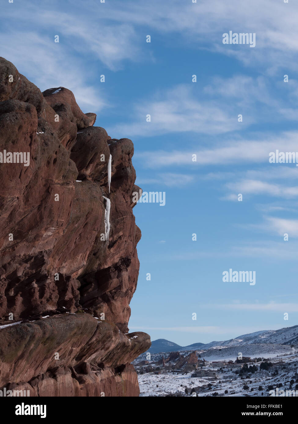 Red rock cliff with icicles and snow, hills in background; rural ...
