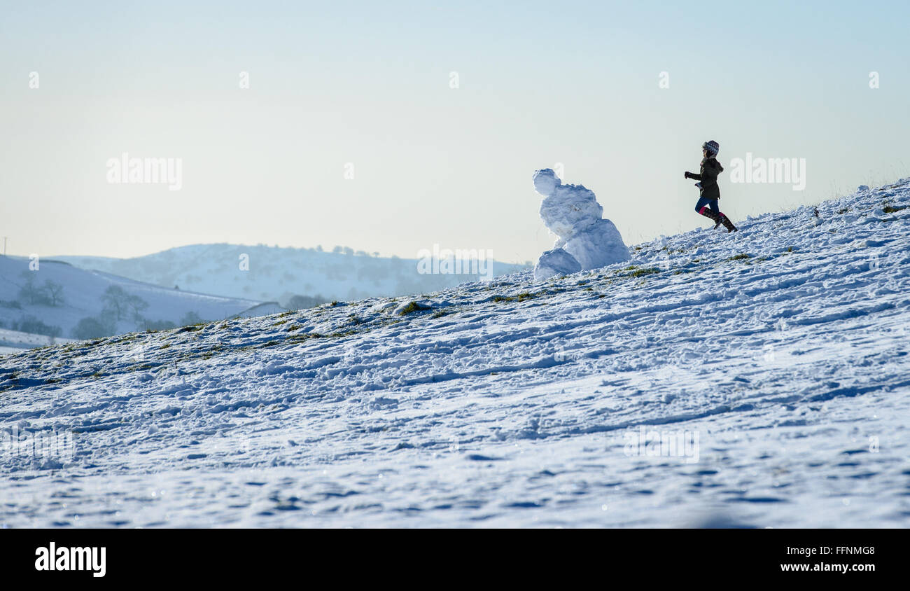 Young girl running down a snow covered hillside, past a leaning snowman ...