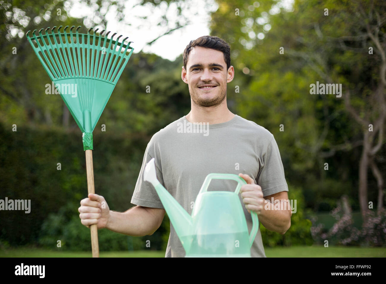 Portrait of young man standing with a gardening rake and watering can ...