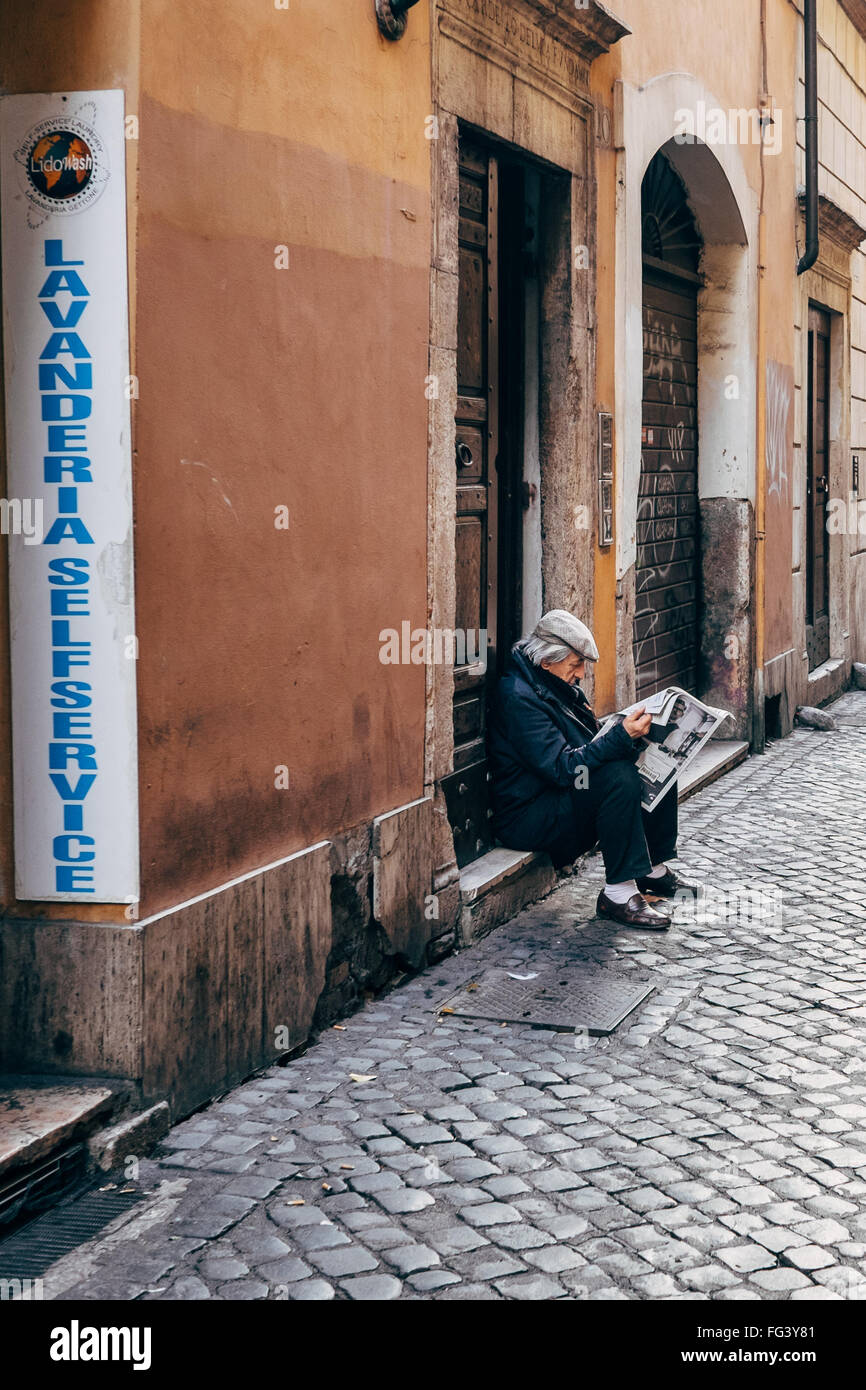 An elederly man reads a newspaper while sitting on a door step in Rome ...