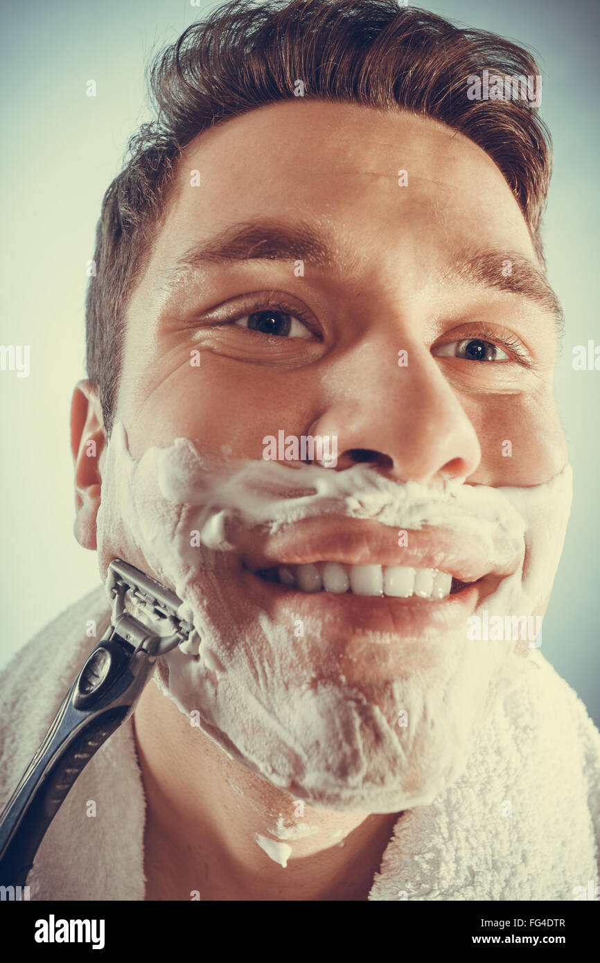 Happy smiling man shaving using razor with cream foam. Handsome guy removing face beard hair ...