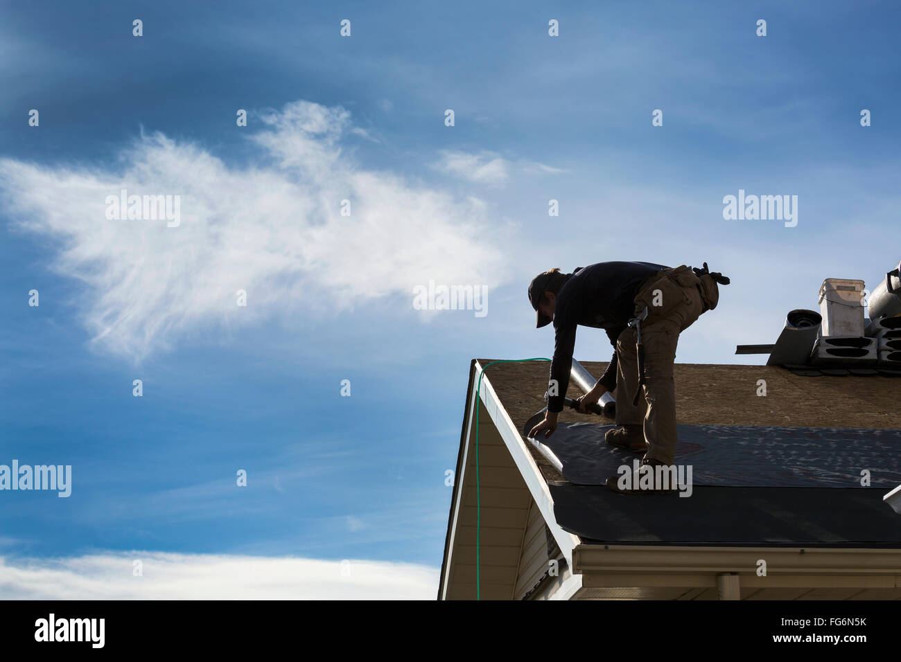 Silhouette of male roofer on top of roof stapling tar paper with clouds ...