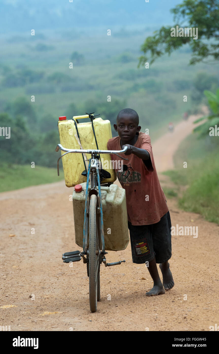 Boy pushing a bike laden with full water containers along a dusty road ...