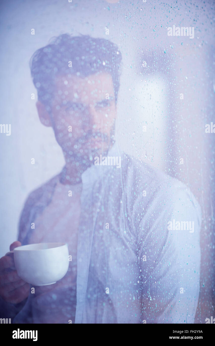 relaxed young man drink first morning coffee withh rain drops on window ...