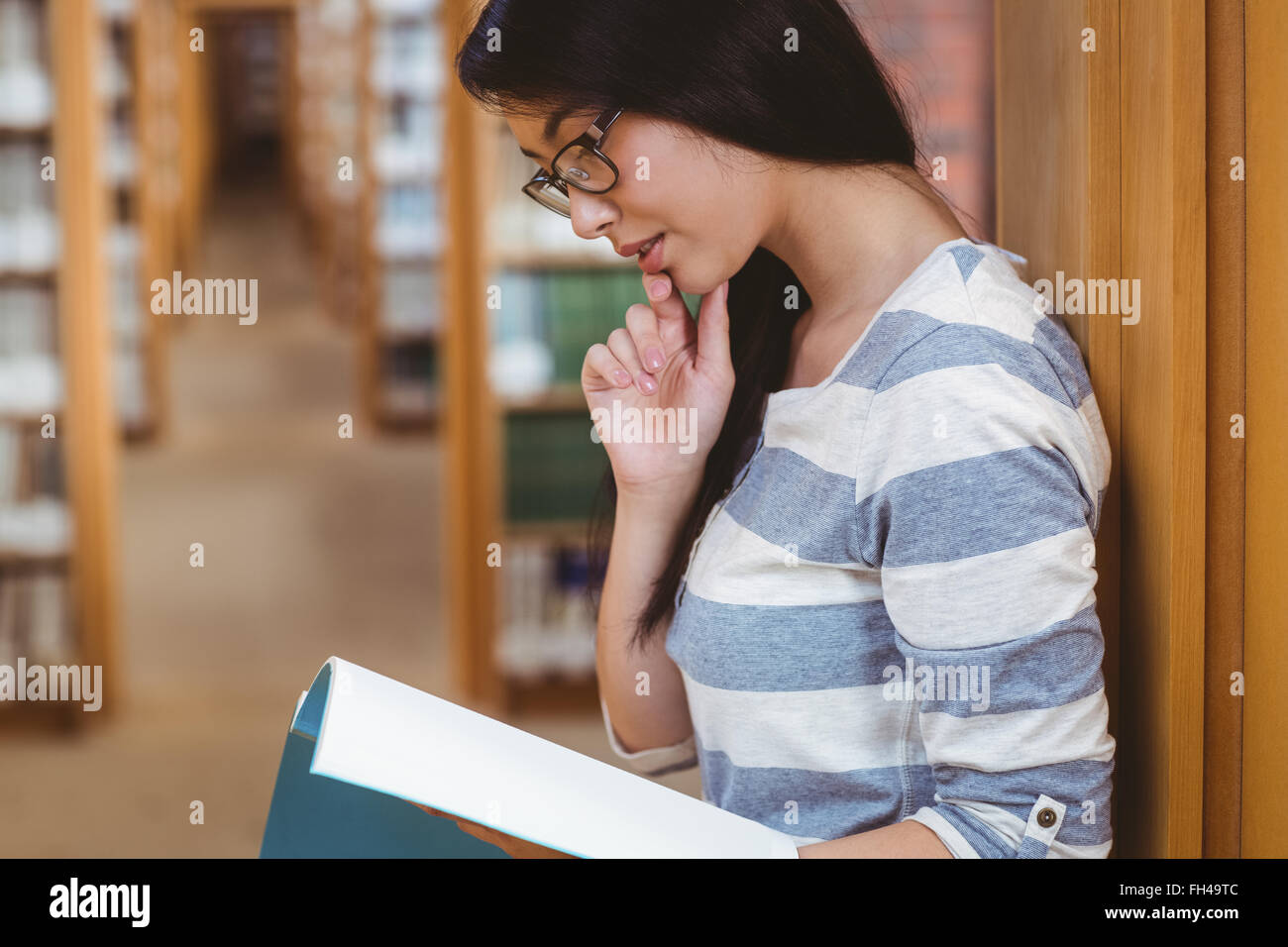Focused student leaning against bookshelves and reading a book in ...