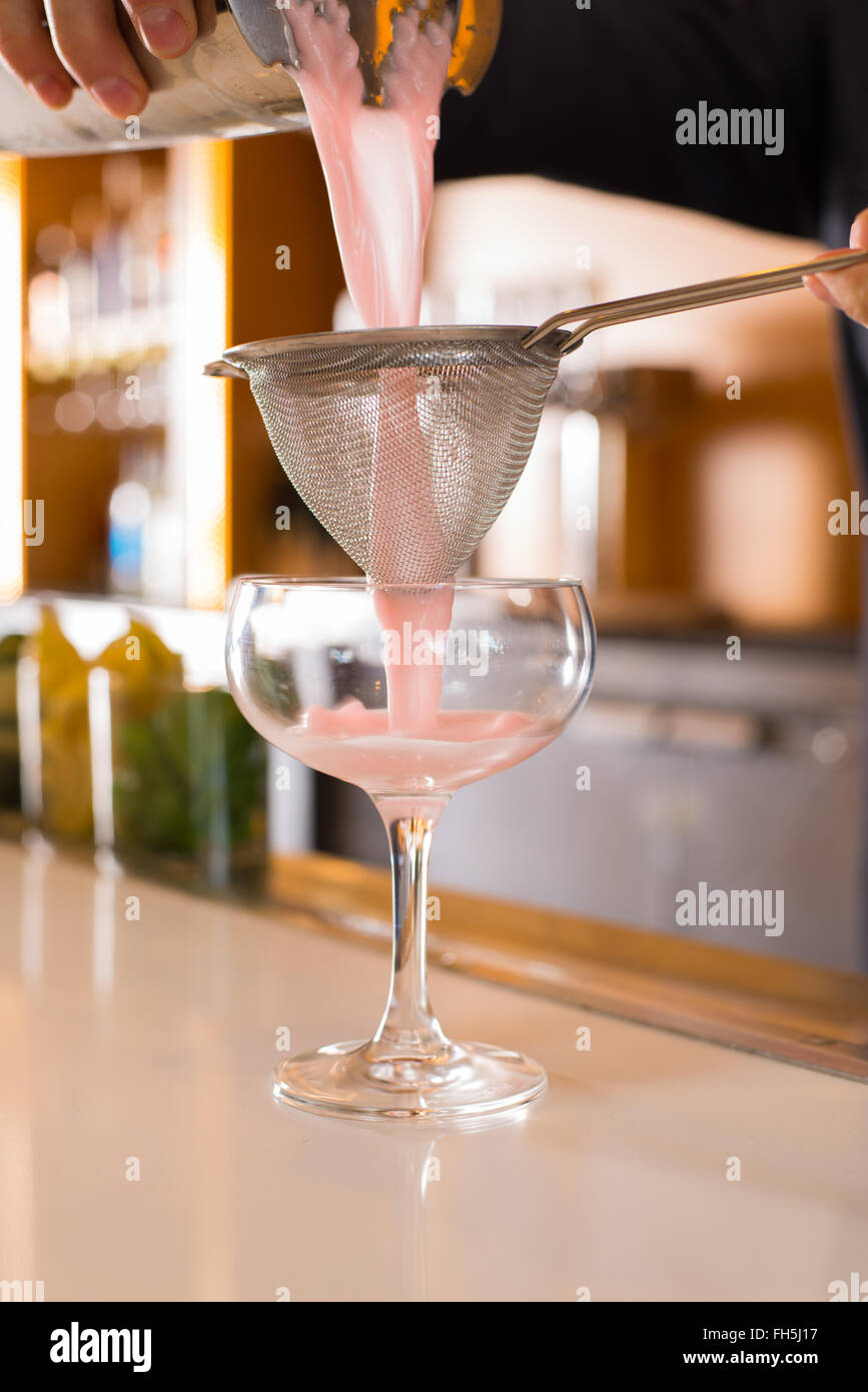 A pink-colored cocktail being poured through a strainer into a stemmed ...