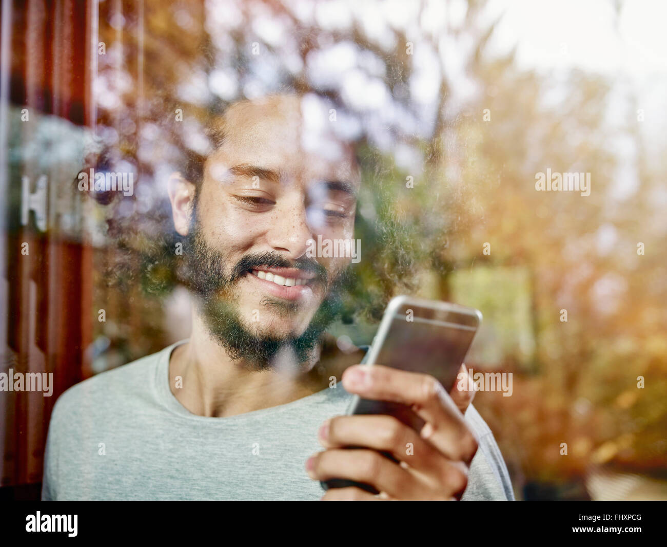 Smiling young man looking on cell phone behind windowpane Stock Photo ...