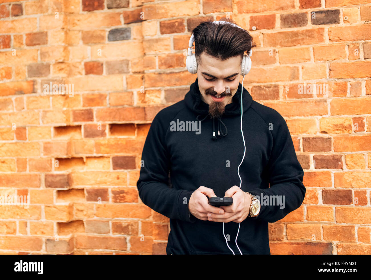 Portrait of young man in front of brick wall listening music with ...